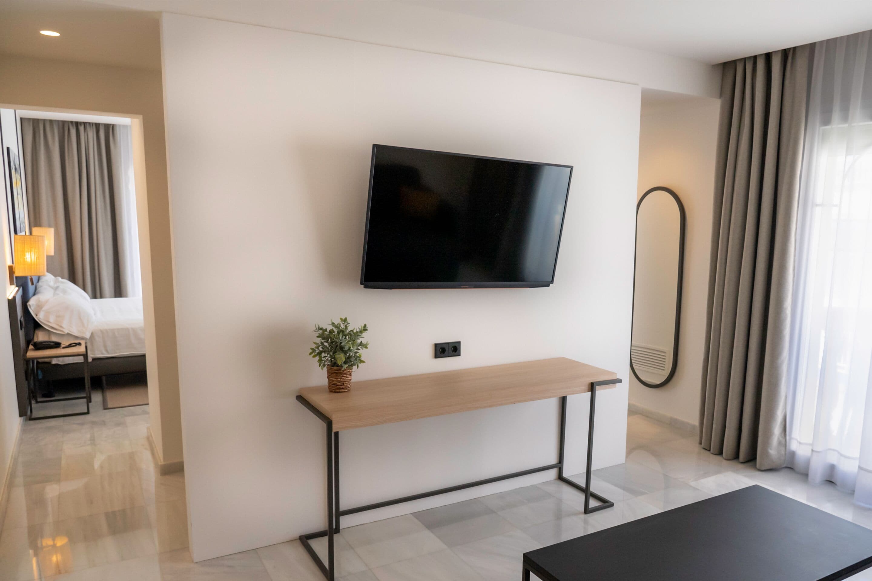 Quarto de hotel com televisão de tela plana na parede, mesa consola de madeira, mesa de centro preta, cama com roupa de cama branca, abajur e um espelho grande.