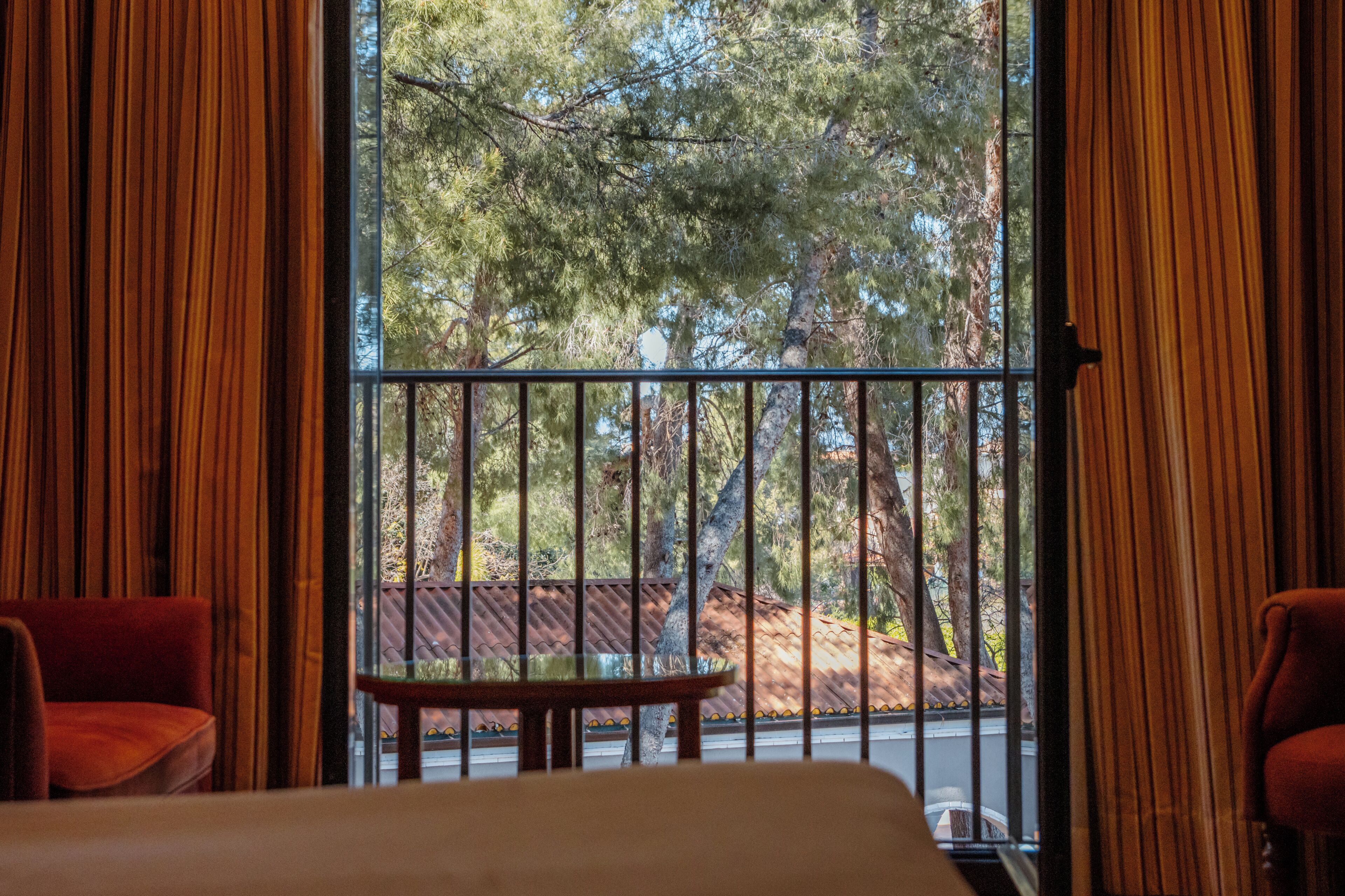 Quarto com cama, uma mesinha e vista para árvores através de uma janela com cortinas.