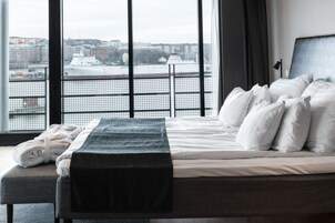Quarto de hotel com uma cama, um banco e vista da cidade através de amplas janelas.