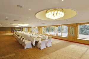 Uma sala de banquetes com longas mesas preparadas para um evento formal, grandes janelas e um lustre.