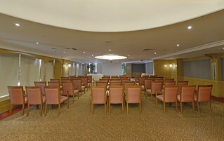 Uma sala de conferência com fileiras de cadeiras cor de rosa, piso carpete e um grande lustre.