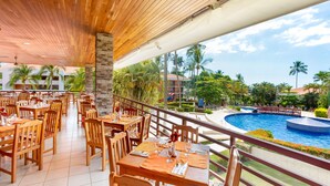 Um restaurante com mesas e cadeiras de madeira, piscina e vista para palmeiras e edifícios.