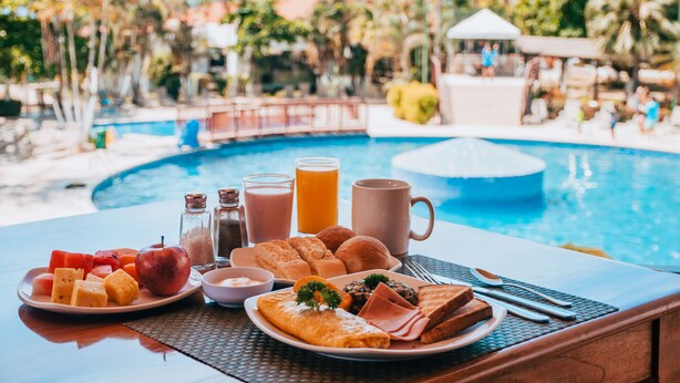 Mesa de pequeno-almoço com diversas opções de comida e um copo de sumo de laranja, com uma piscina e palmeiras ao fundo.