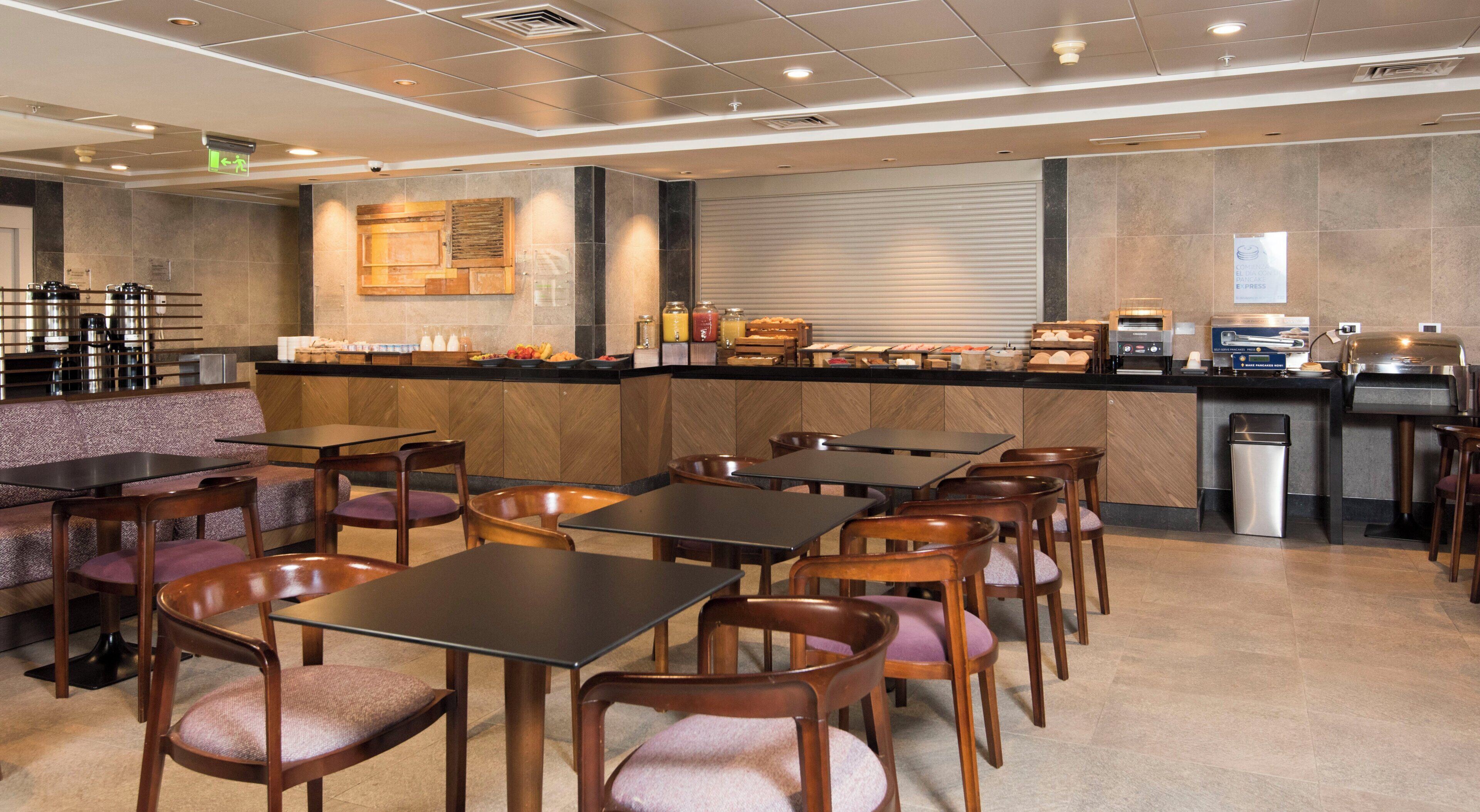 Uma área de refeições de hotel com mesas e cadeiras, um balcão com comida e bebidas, e uma máquina de café.
