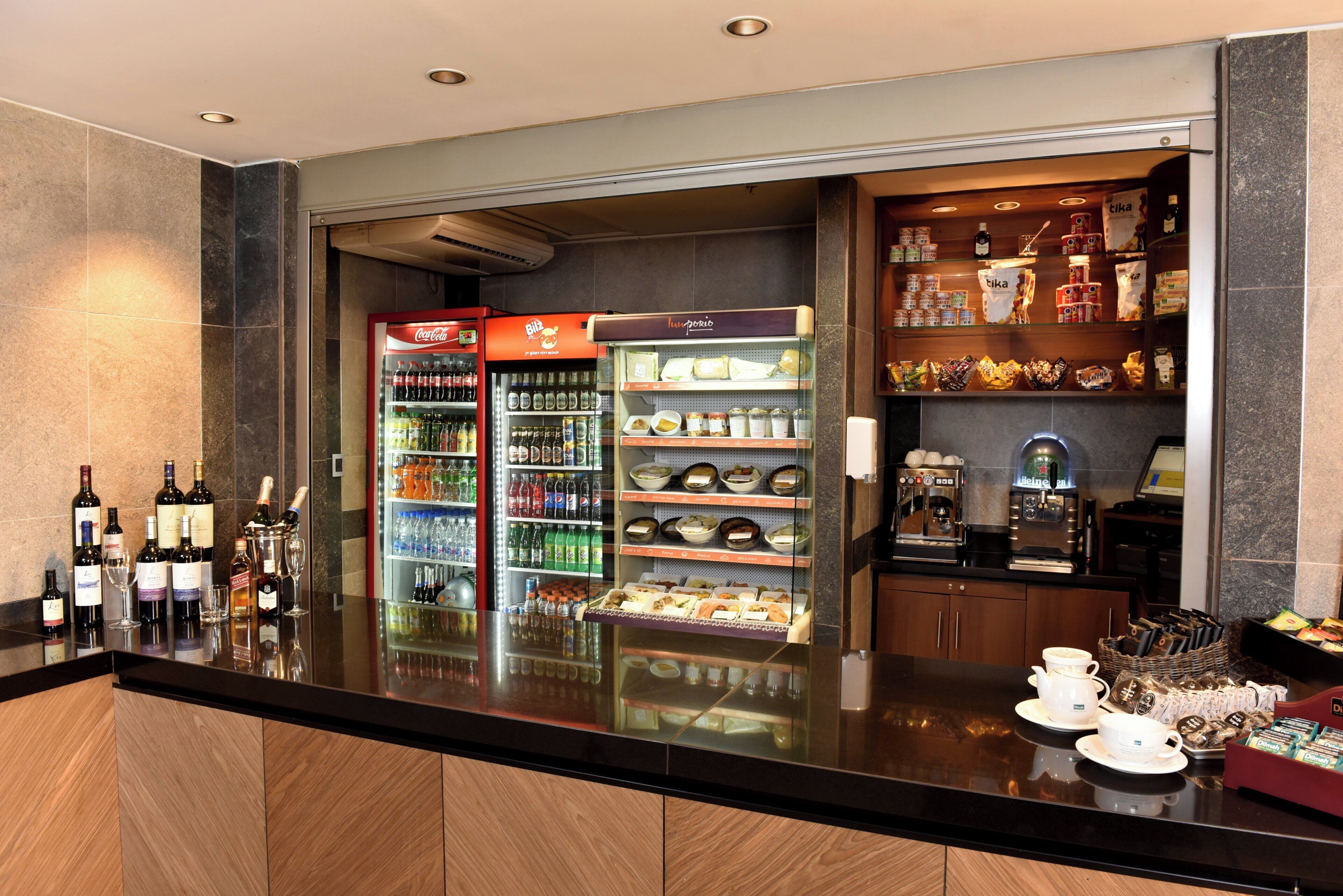 Um saguão de hotel com balcão de recepção, refrigerador Coca-Cola, exposição de bebidas geladas e máquina de café.