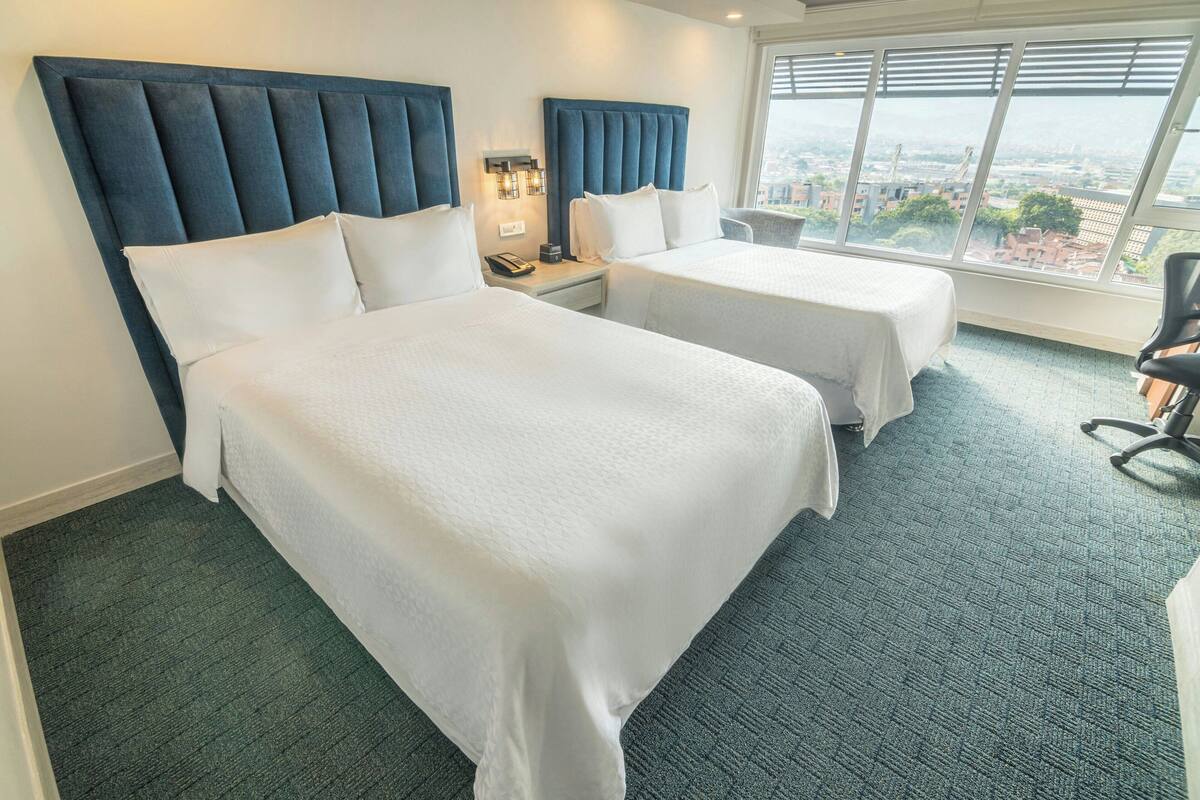 Quarto de hotel com duas camas, uma mesa de cabeceira com abajur, uma cadeira e uma janela grande com vista para a cidade.