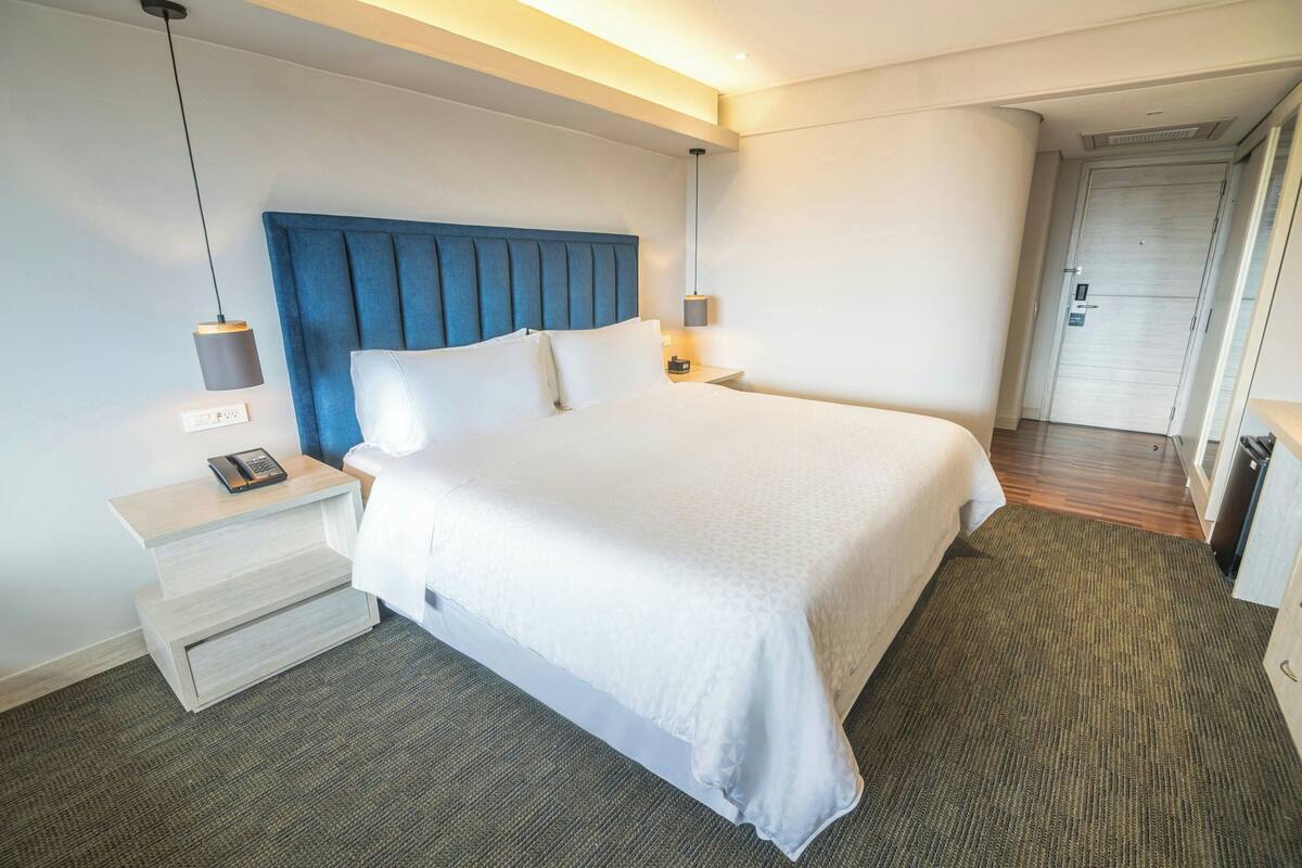 Quarto de hotel moderno com uma cama grande, mesa de cabeceira com abajur, telefone e pequena gaveta.