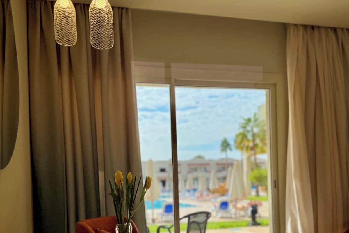 Quarto moderno com uma mesa de madeira redonda, duas cadeiras laranja, um vaso com flores amarelas e vista para uma piscina e palmeiras através de grandes portas de vidro deslizantes.