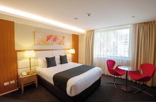 Quarto de hotel com uma cama grande, duas luminárias de cabeceira, uma mesinha com duas cadeiras vermelhas e uma janela com cortinas.