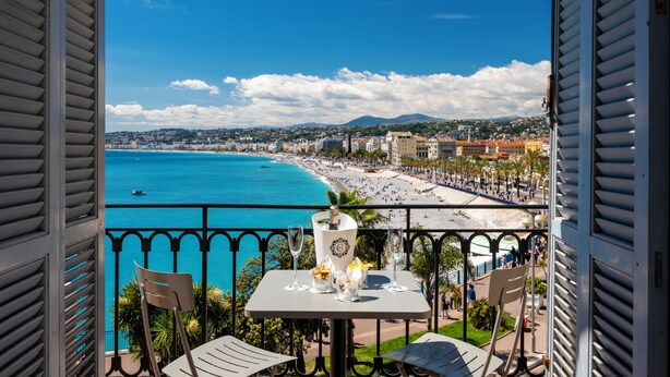 Uma varanda com mesa posta para dois, com vista para uma praia e um mar azul cristalino, além de um horizonte urbano ao fundo.