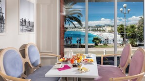 Uma área de refeições com mesa posta para o café da manhã, com vista para uma praia com pessoas e palmeiras.