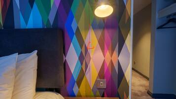 Quarto de hotel com uma cama, parede com padrão de losangos coloridos e luminária pendente.
