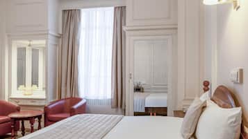 Quarto de hotel com uma cama grande, área de estar com mesa e cadeiras, toucador com espelho e uma janela com cortinas.