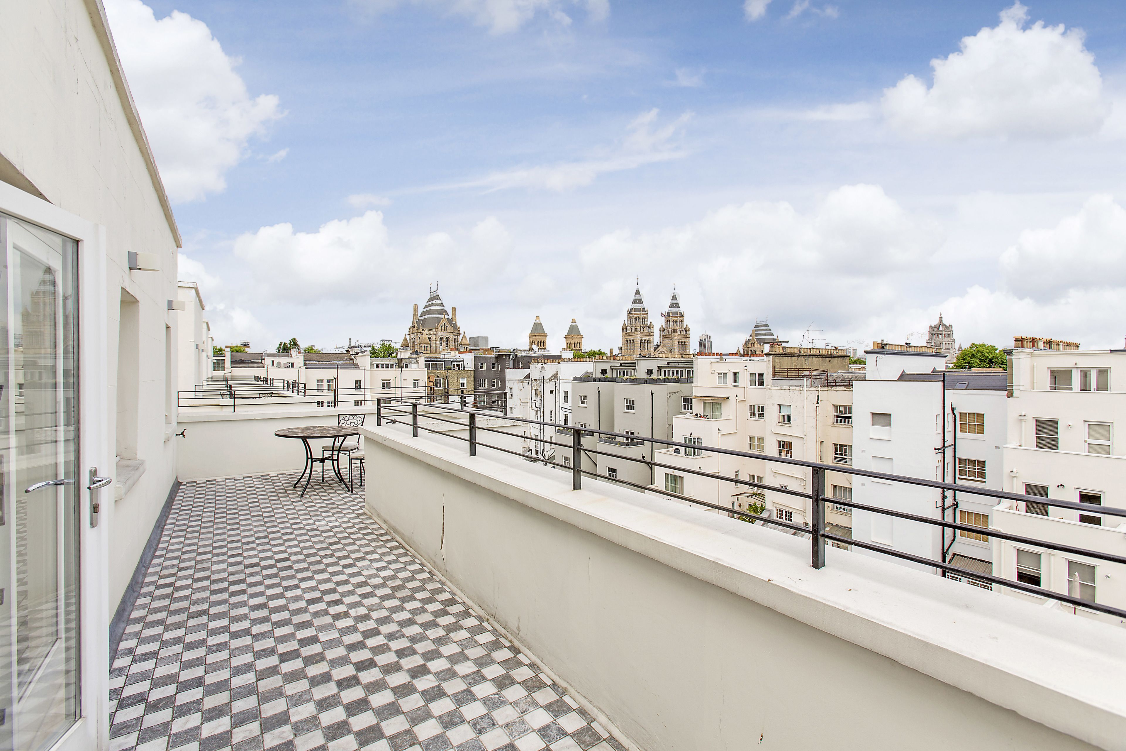 Uma varanda com piso xadrez, uma mesa e cadeiras, com vista para um horizonte urbano com edifícios históricos.