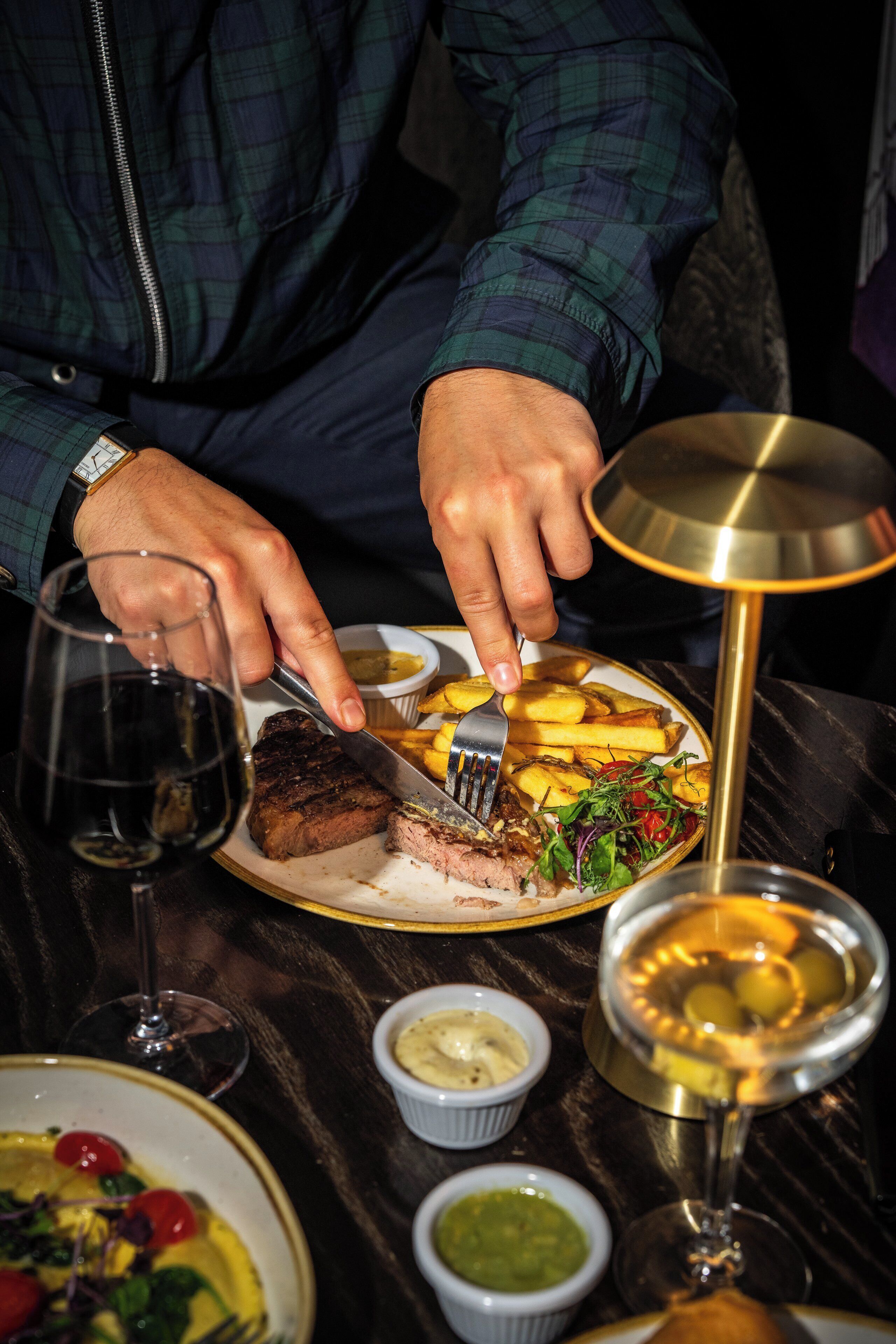 Uma pessoa jantando à mesa, com um bife, batatas fritas e salada, além de uma taça de vinho e pequenos recipientes com acompanhamentos.