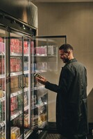 Uma pessoa vestindo uma camisa escura em frente a uma vitrine refrigerada com diversos alimentos.