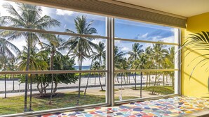 Quarto com uma grande janela com vista para uma praia, palmeiras e céu claro.