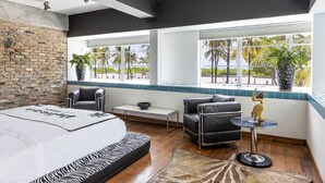 Quarto de hotel moderno com uma cama grande, área de estar com mesa, e vista para palmeiras através de janelas amplas.