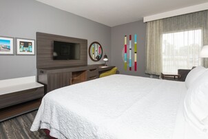 Quarto de hotel com uma cama grande, televisão de tela plana fixada em um painel de madeira, escrivaninha com cadeira, mesa pequena com abajur e janela com cortinas finas.