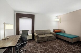 Quarto de hotel com uma mesa de jantar, um sofá, uma cadeira, um abajur e uma janela.