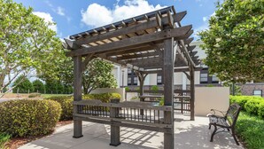 Uma pérgola de madeira com um banco por baixo, rodeada por vegetação e um edifício ao fundo.