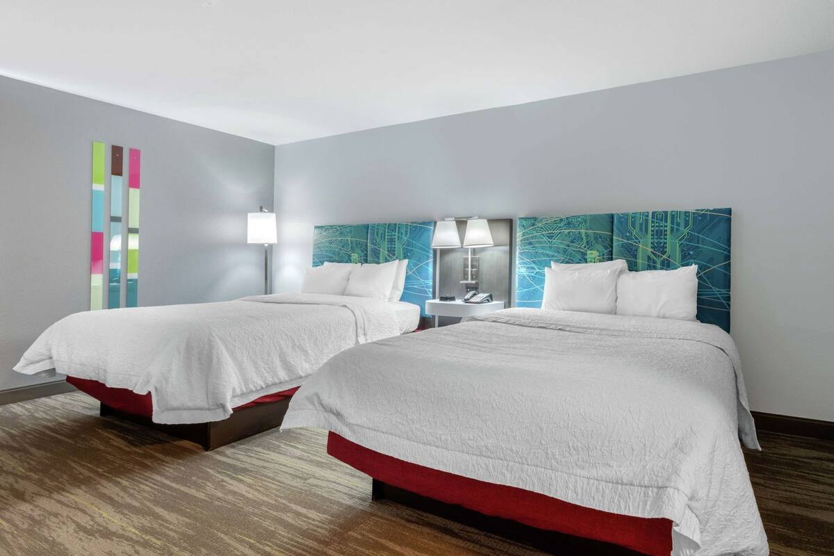 Quarto de hotel com duas camas, uma mesa de cabeceira com abajur e uma parede com padrão geométrico.