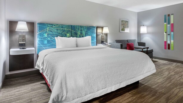 Quarto de hotel moderno com uma cama grande, mesas de cabeceira com abajures, uma escrivaninha e uma obra de arte abstrata colorida na parede.