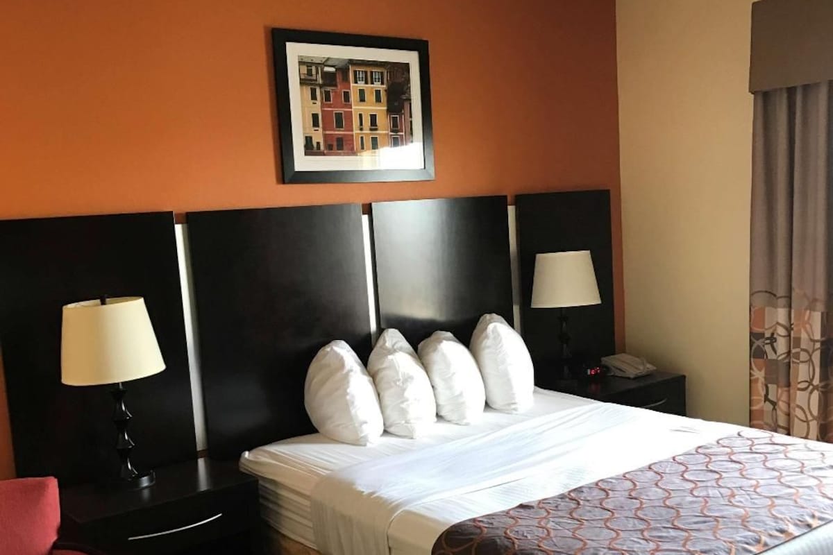 Quarto de hotel com uma cama grande, duas mesas de cabeceira com abajures, uma poltrona vermelha e um quadro na parede.