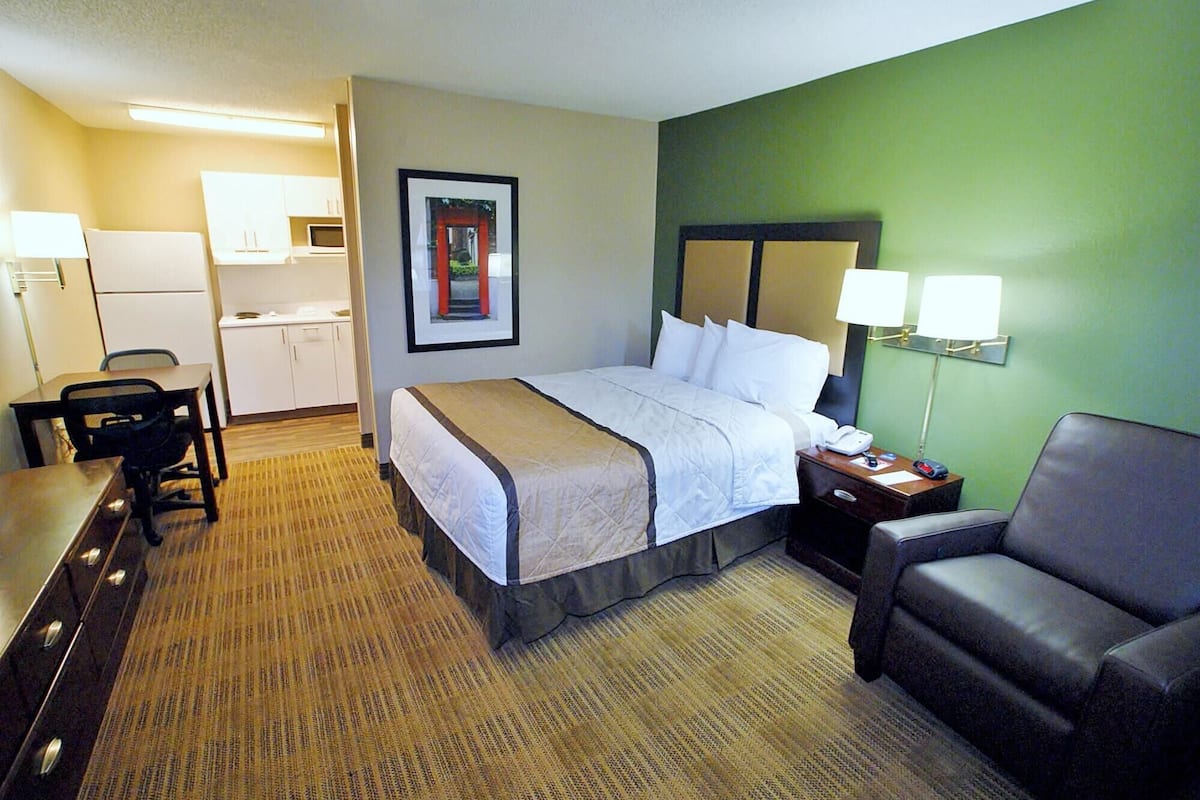Quarto de hotel com cama, escrivaninha, cadeira, mesa de cabeceira e um quadro na parede.