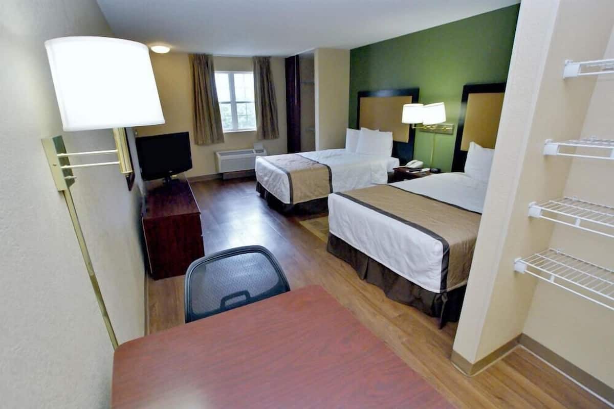 Quarto de hotel com duas camas, uma mesa, uma cadeira, uma TV e um abajur.