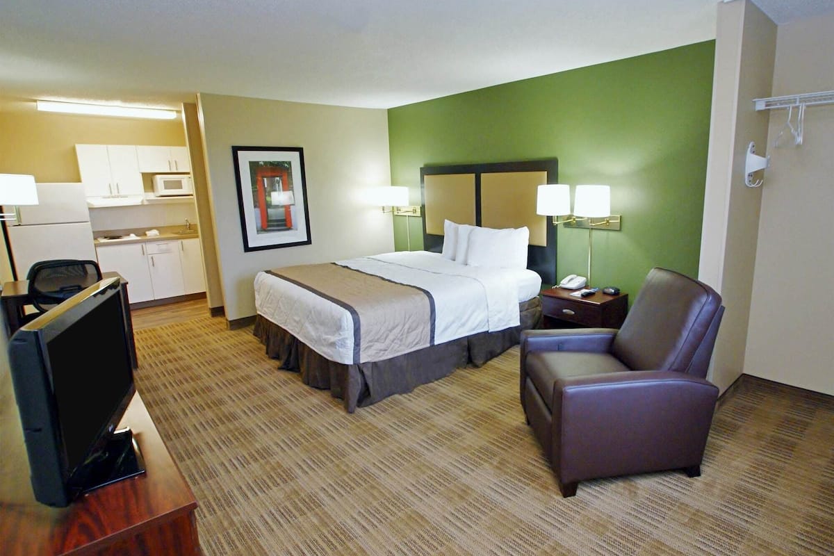 Quarto de hotel com cama, escrivaninha com computador, cadeira, mesa de cabeceira com luminárias e uma parede verde decorativa.