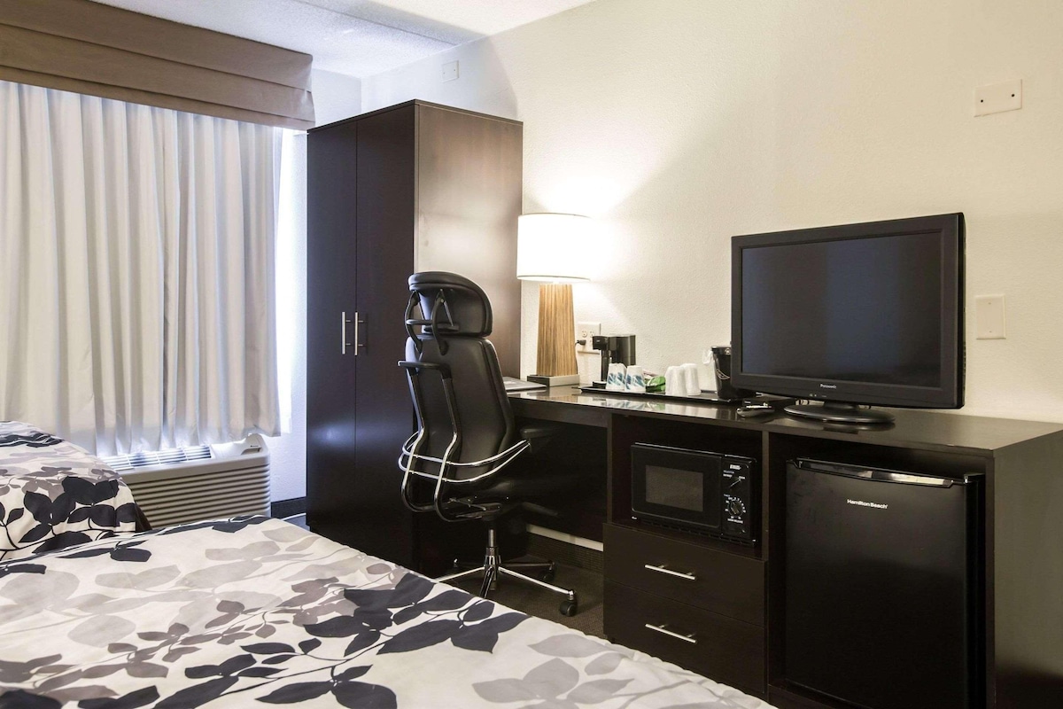 Quarto de hotel com cama, escrivaninha com televisão, cadeira, luminária e minibar.