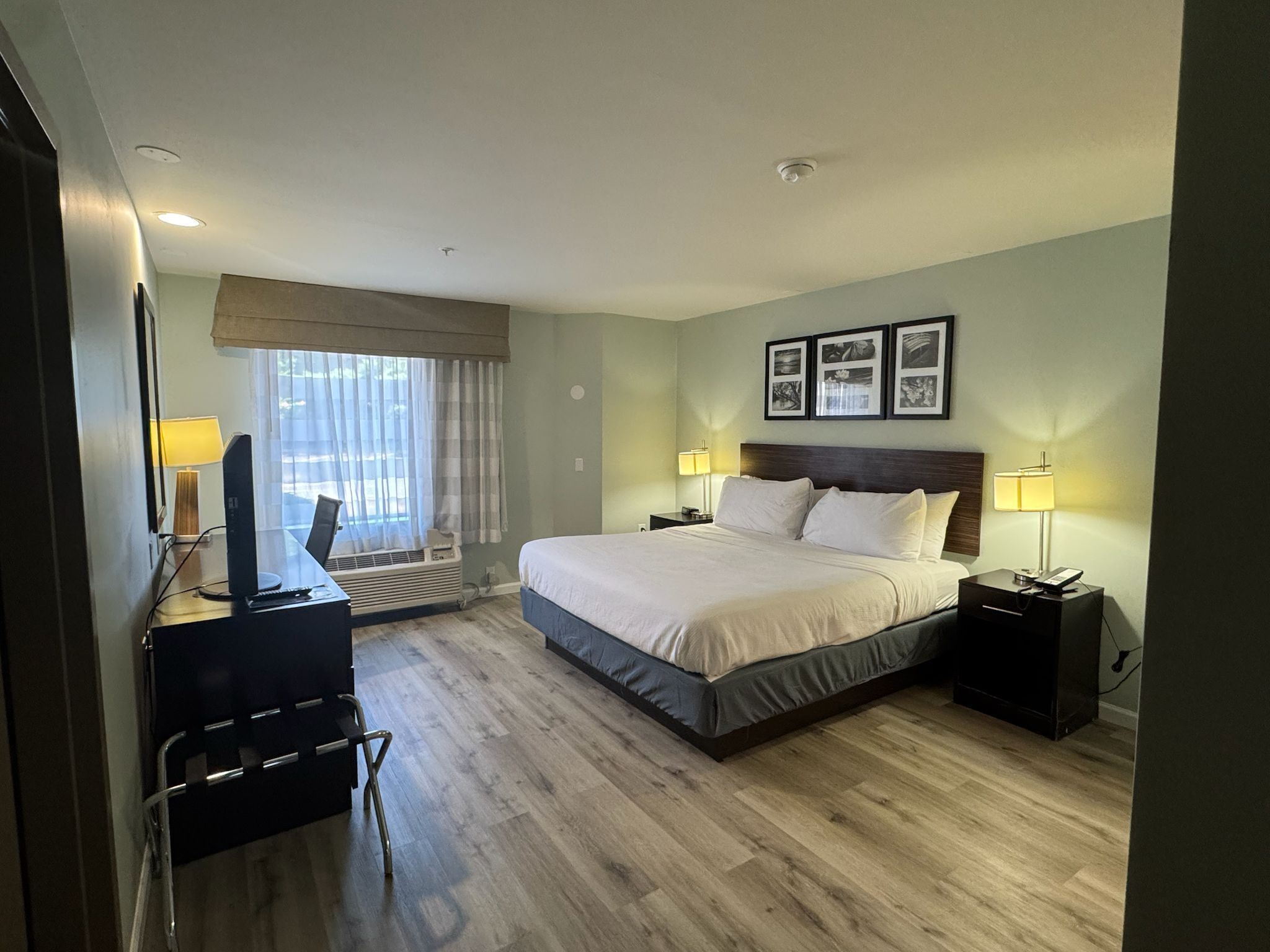 Quarto de hotel com cama, mesas de cabeceira, escrivaninha e computador, e janela com cortinas.