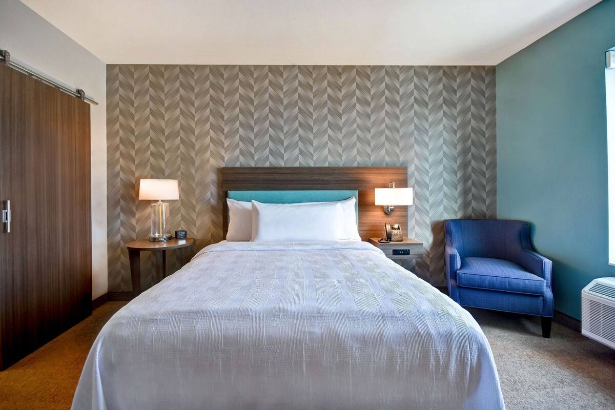 Quarto de hotel com uma cama grande, duas mesas de cabeceira com luminárias, uma poltrona azul e um guarda-roupa.