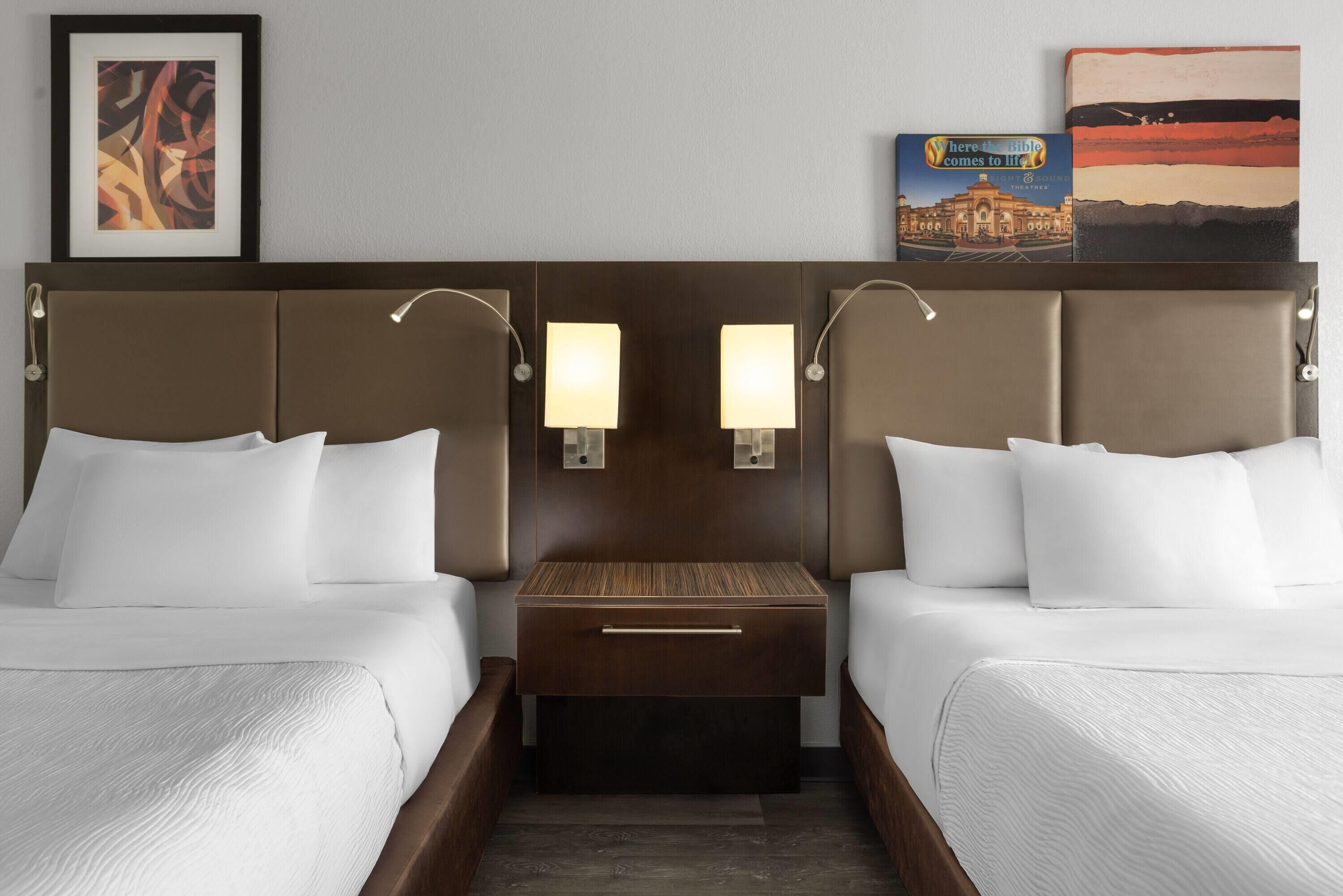 Quarto de hotel com duas camas, uma mesa de cabeceira e luminárias fixadas na parede.