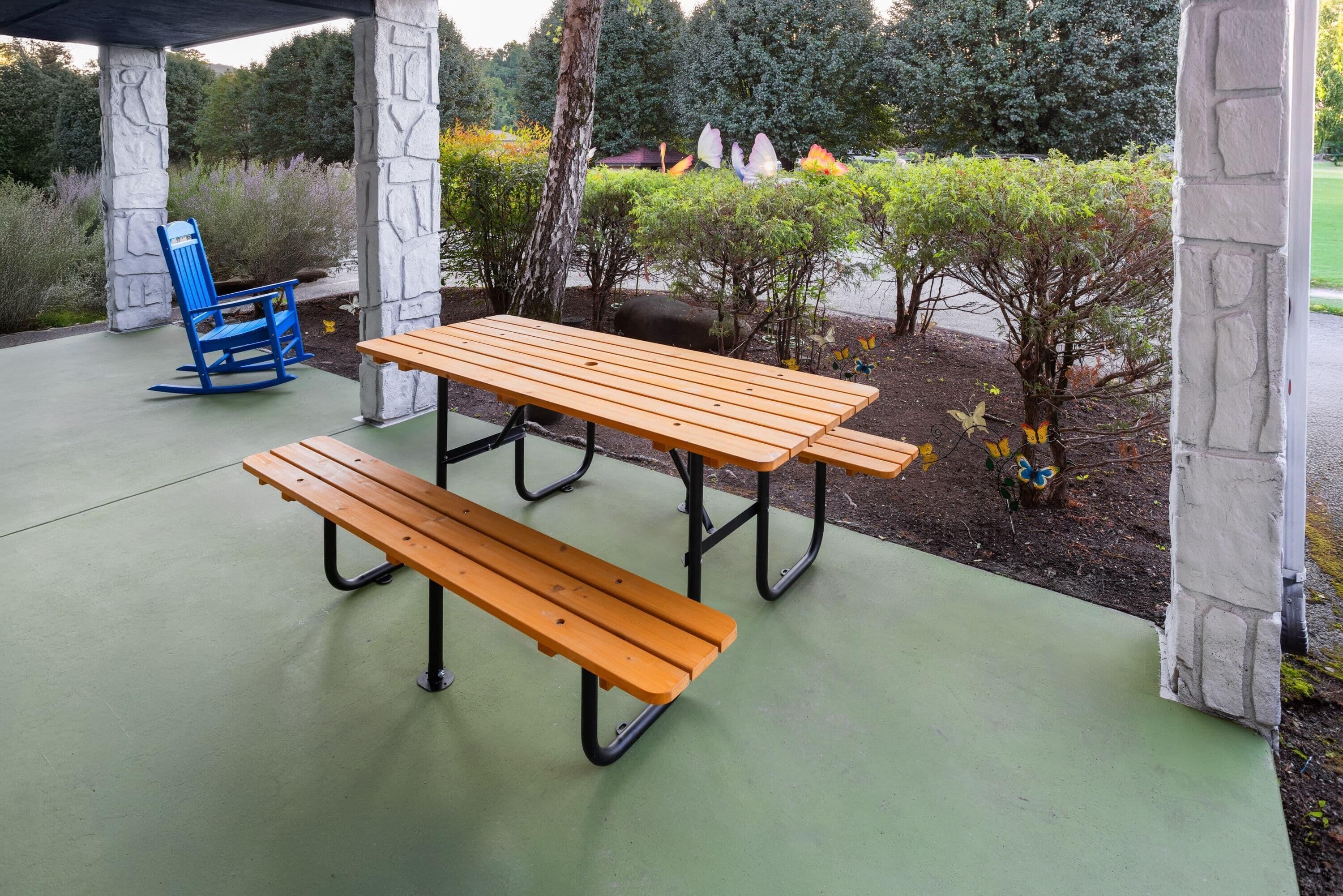 Um pátio externo com mesa e bancos de madeira, rodeado por pilares de pedra e vegetação.