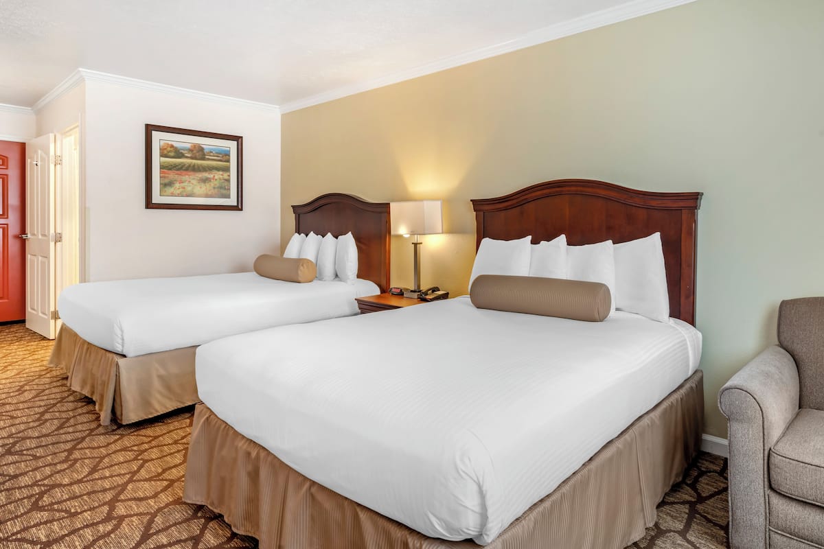 Quarto de hotel com duas camas, uma mesa de cabeceira, uma luminária e uma cadeira.