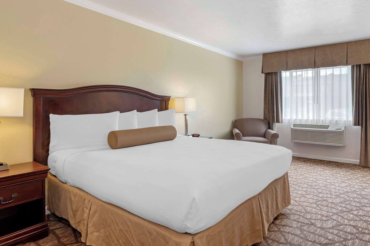 Quarto de hotel com uma cama grande, duas luminárias laterais, uma cadeira e um aparelho de ar condicionado.