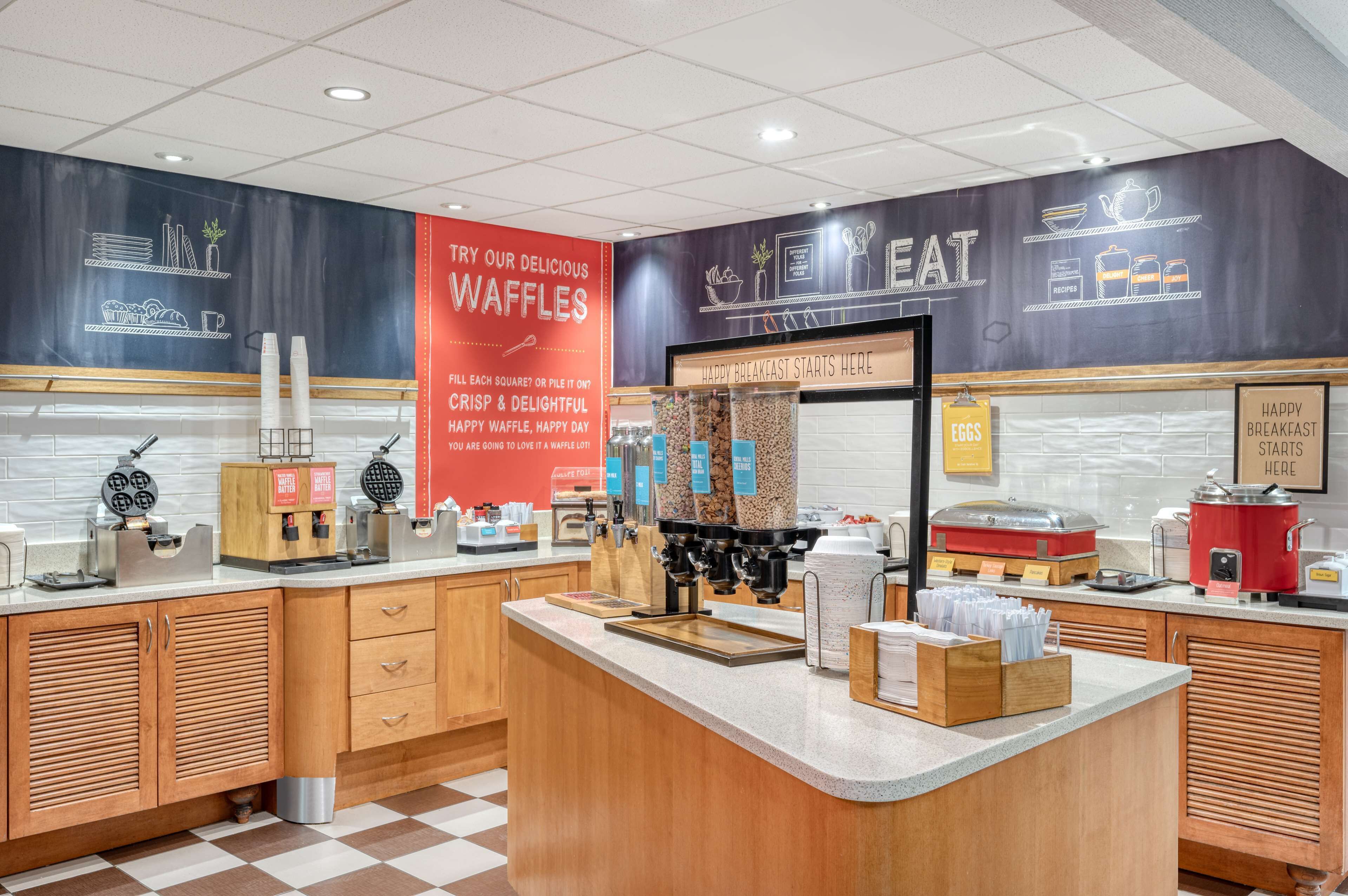 Espaço de café da manhã com estações de waffles, máquinas de café e um letreiro anunciando waffles.