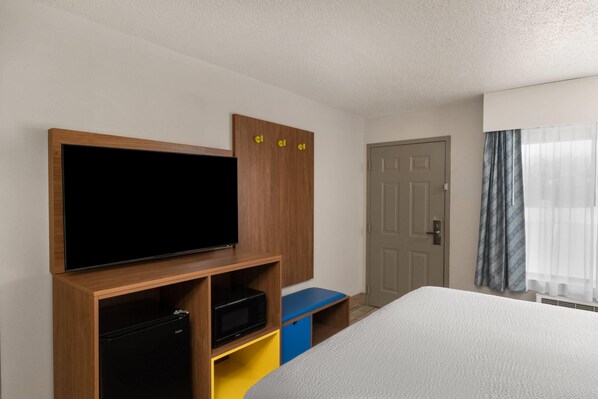 Quarto de hotel com uma cama, uma televisão sobre um suporte de madeira, uma porta e uma janela com cortinas.