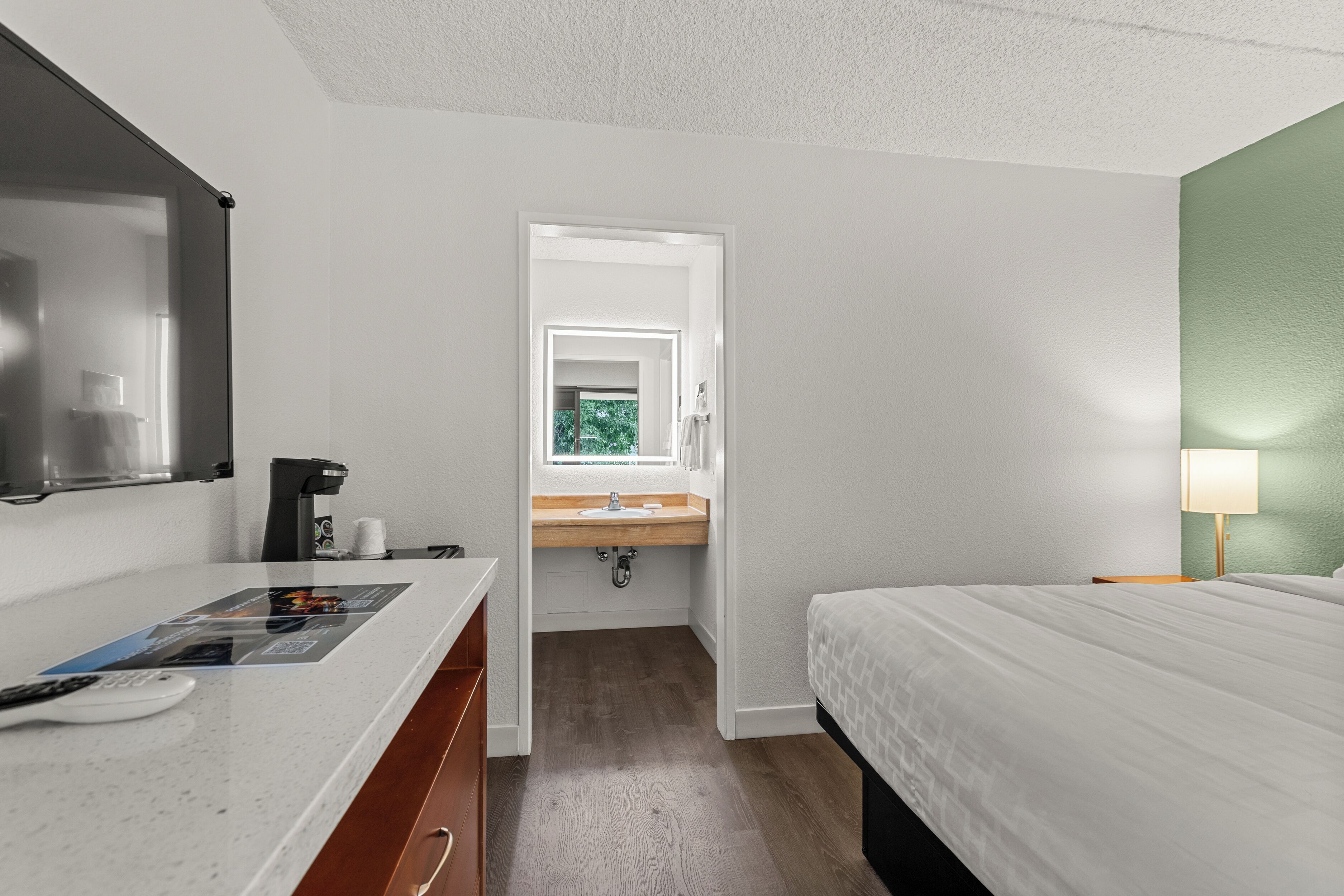 Quarto de hotel com cama, escrivaninha com controle remoto, cafeteira e banheiro com pia e espelho.