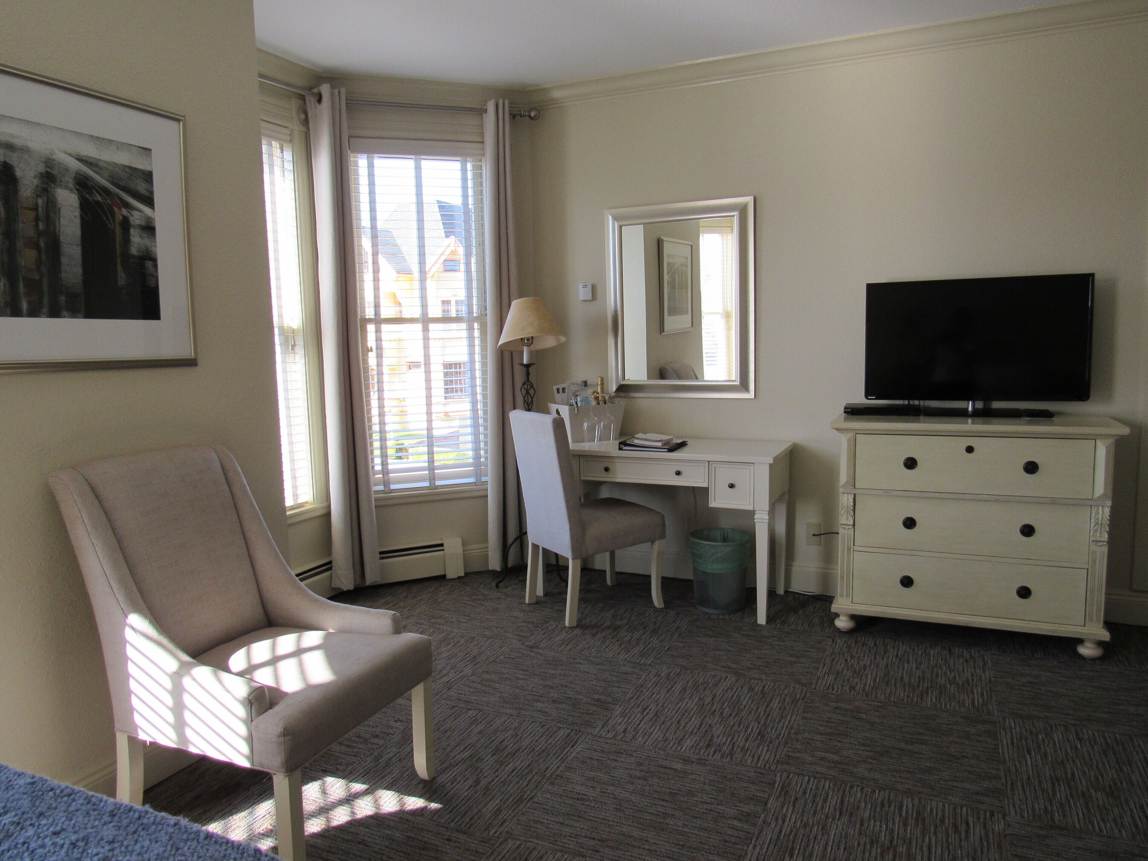 Quarto de hotel com escrivaninha, cadeira, televisão, cômoda e um quadro pendurado na parede.