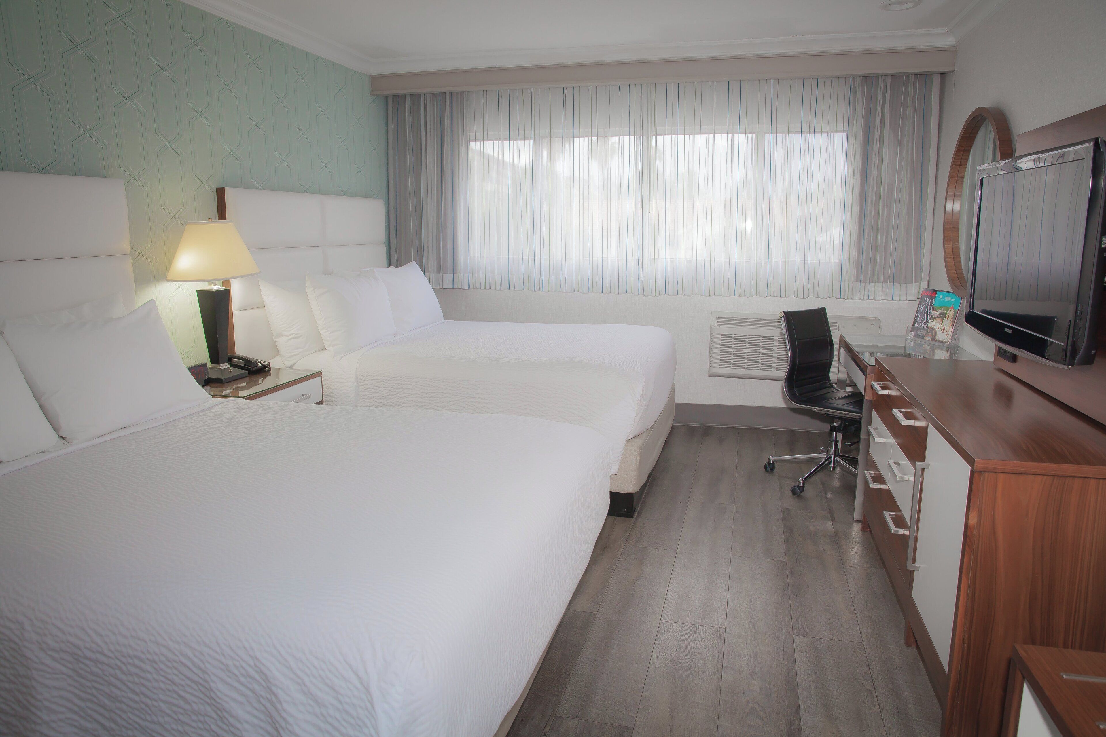 Quarto de hotel com duas camas, uma escrivaninha com televisão, uma cadeira e uma janela com cortinas.