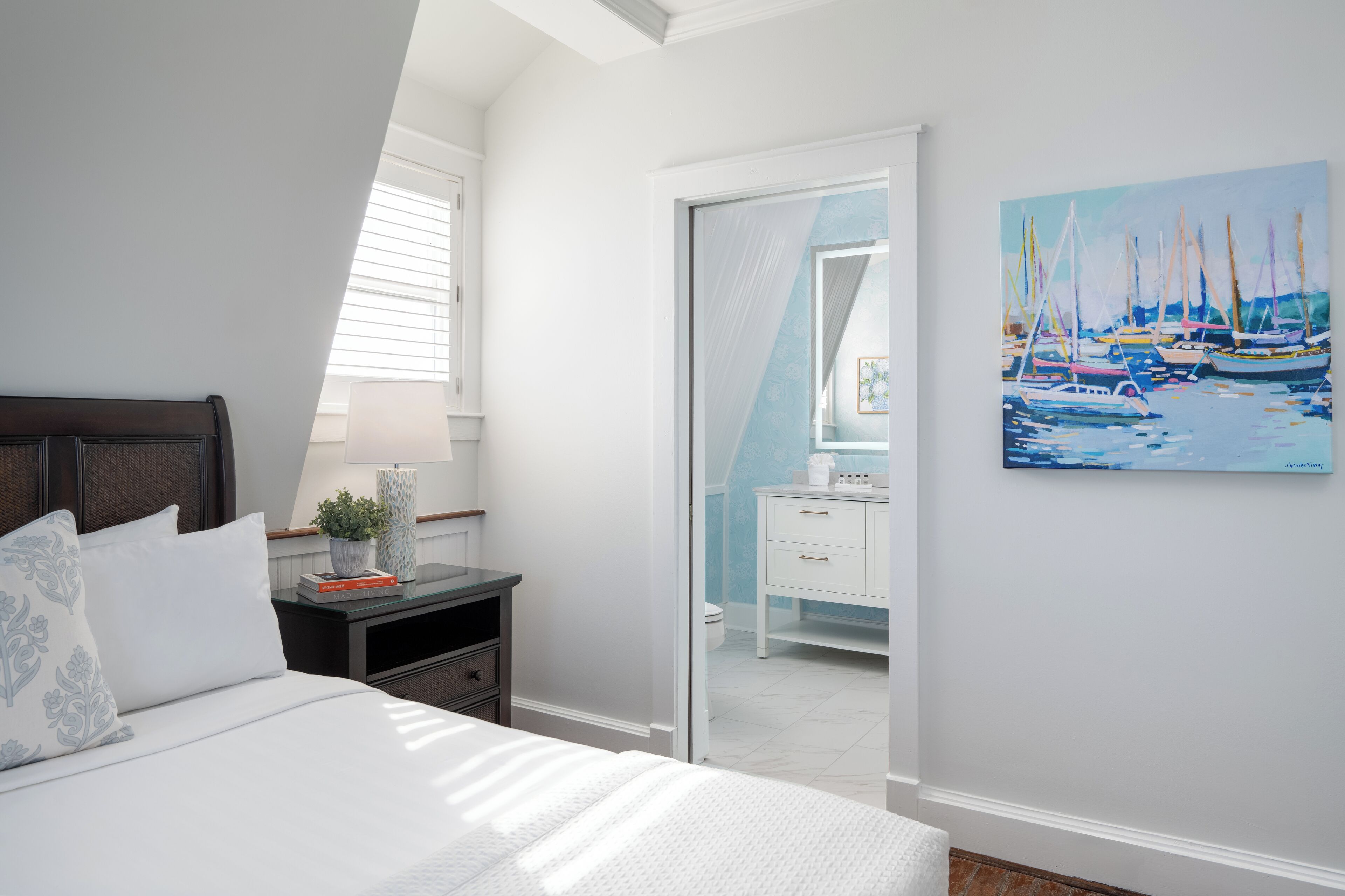 Quarto com cama, mesa de cabeceira, abajur e um quadro de barcos na parede.