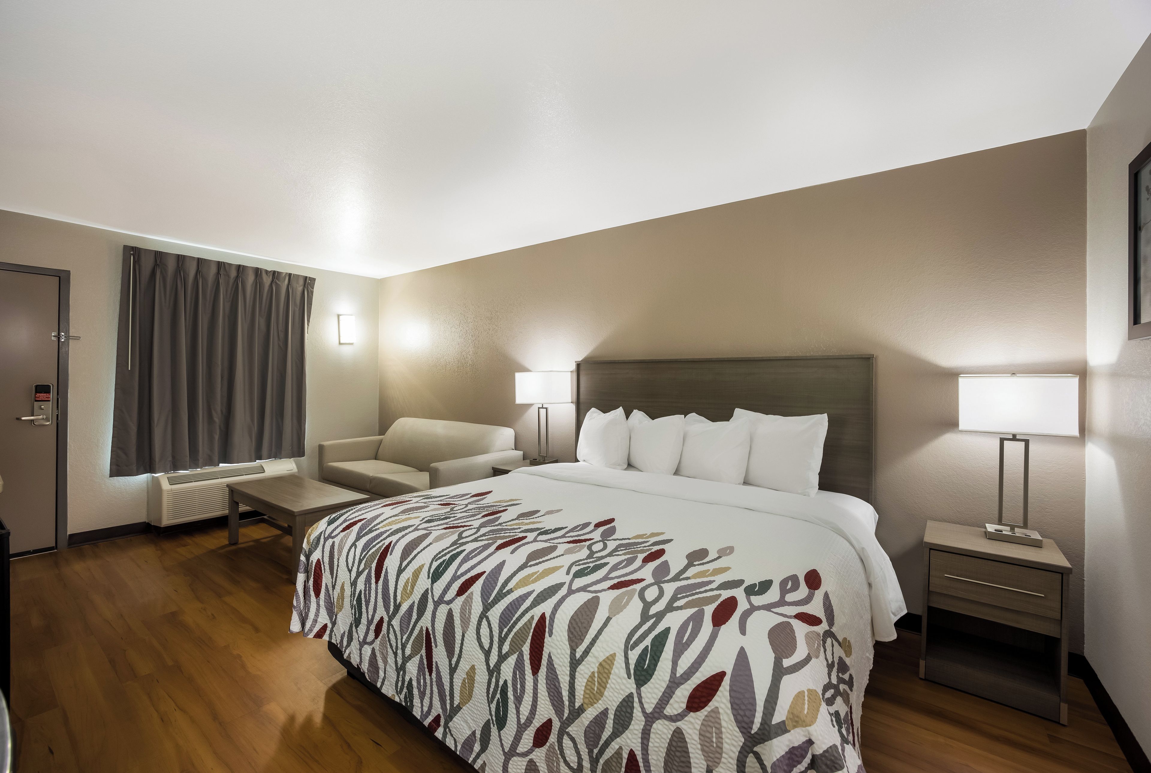 Quarto de hotel com uma cama grande, duas mesas de cabeceira, uma cadeira, uma escrivaninha, um abajur e uma janela com cortinas.