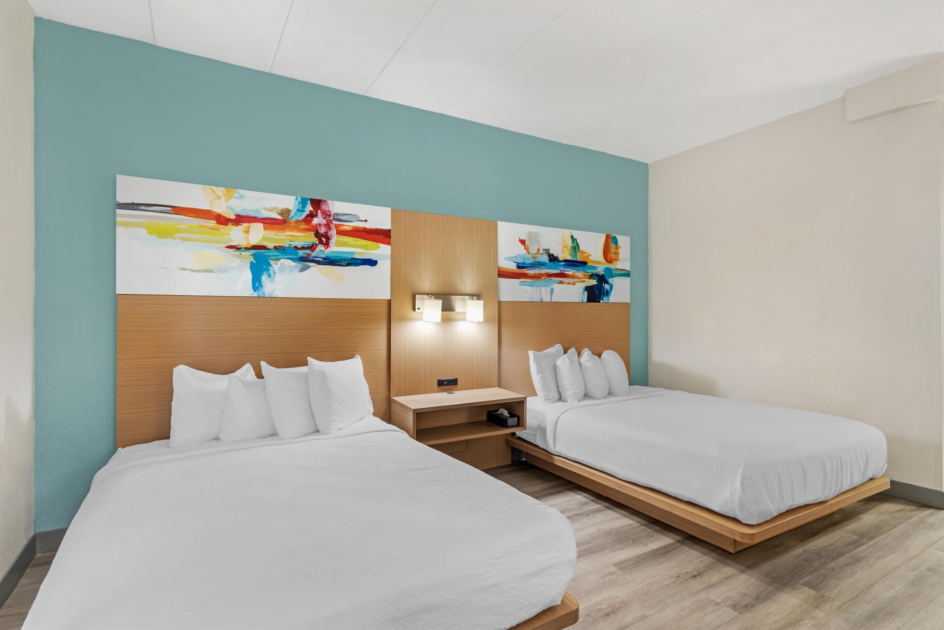 Quarto de hotel com duas camas, cabeceiras de madeira, uma mesa de cabeceira e arte abstrata na parede.