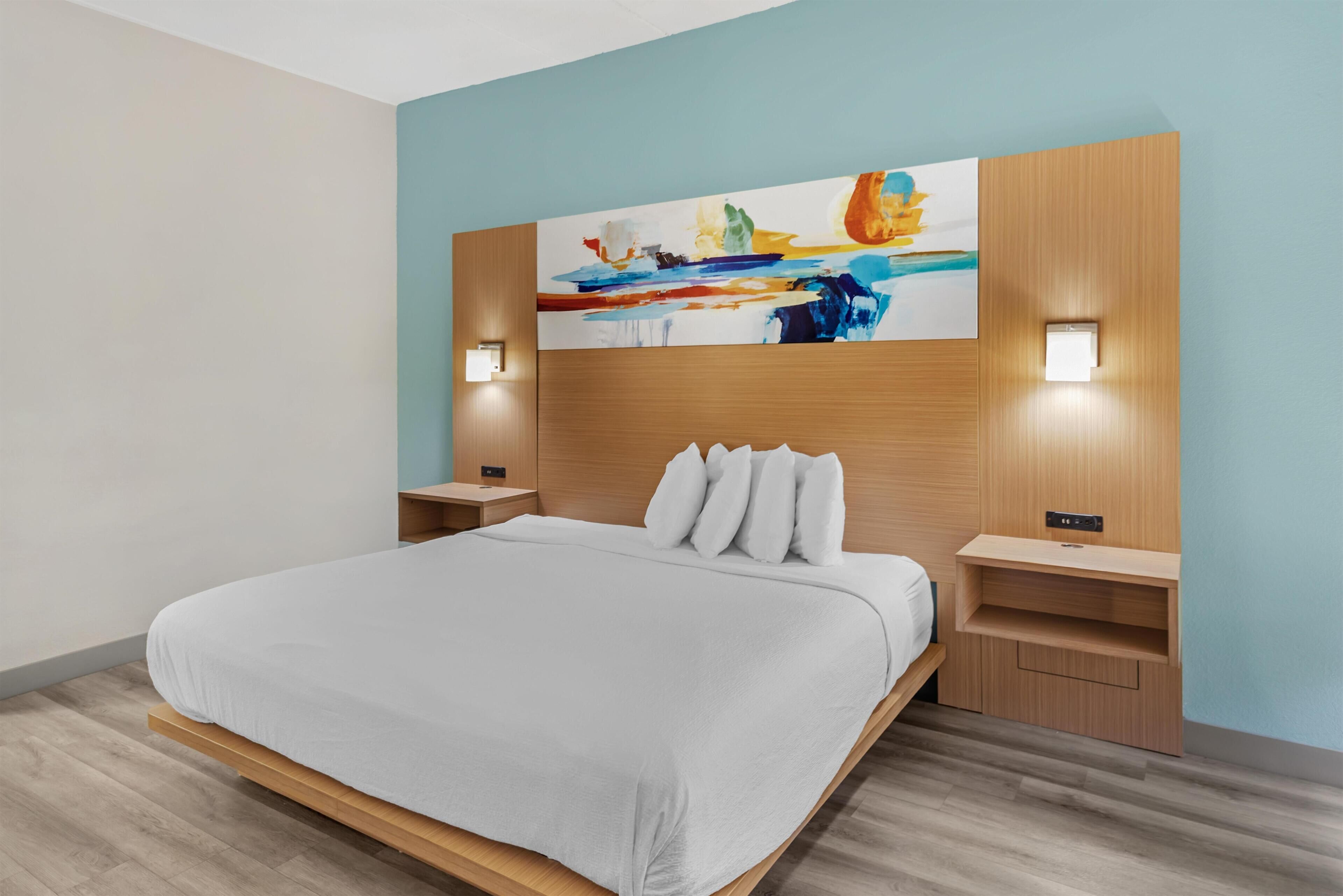 Quarto de hotel moderno com uma cama grande, cabeceira de madeira e uma pintura abstrata colorida sobre a cama.