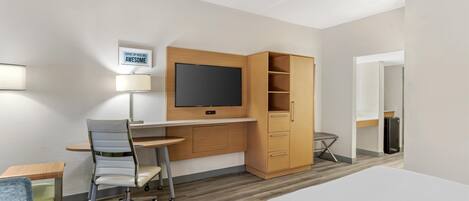 Quarto de hotel com uma cama, uma escrivaninha com cadeira, televisão, guarda-roupa e uma mesinha.