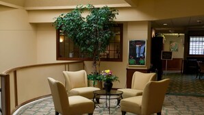 Um lobby com uma escadaria curva, dois poltronas bege, uma mesa redonda com um vaso de flores e uma grande planta em vaso.
