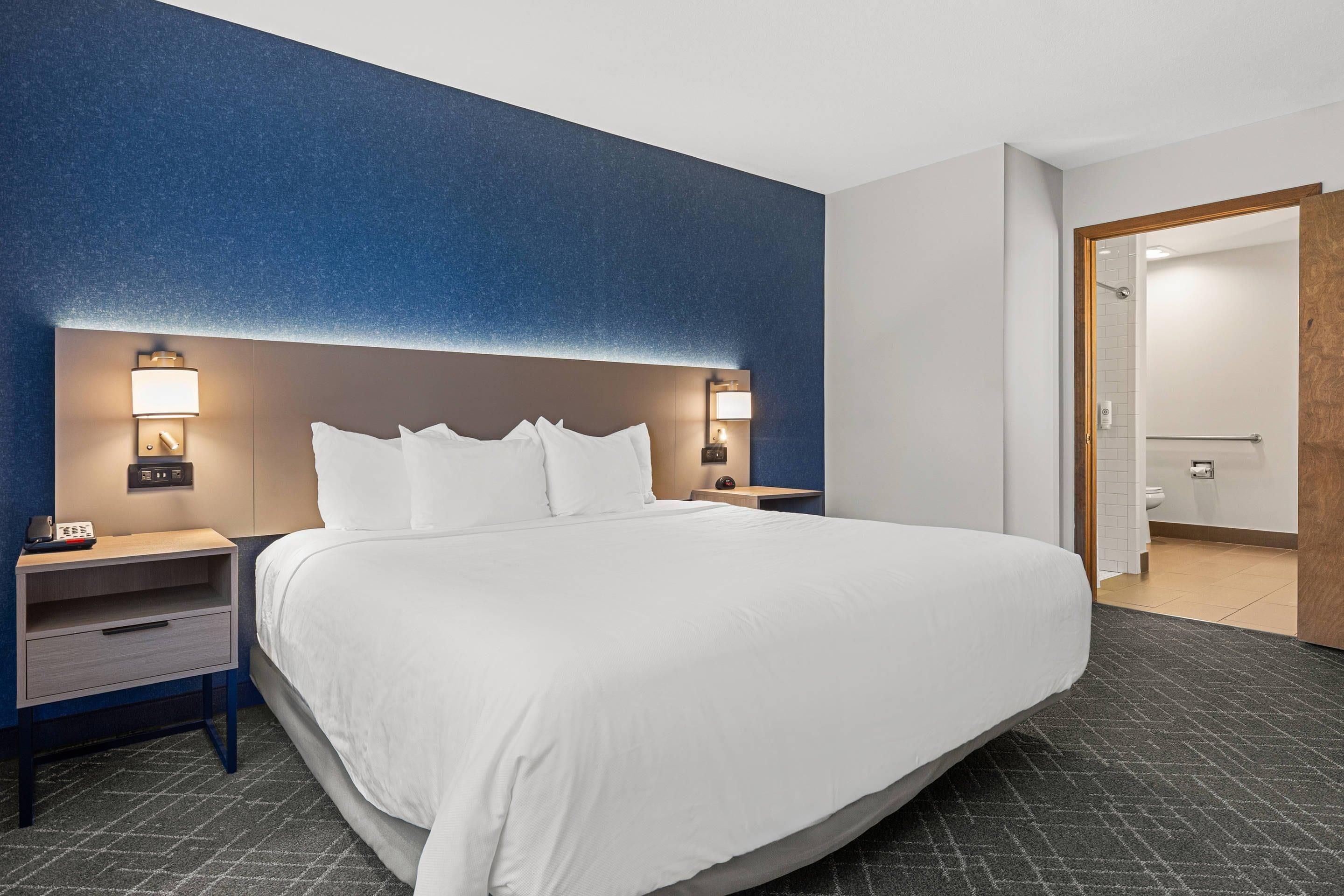 Quarto de hotel com uma cama grande, luminárias de cabeceira, mesa de cabeceira, porta de madeira e banheiro visível através de uma porta aberta.